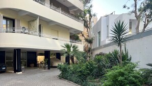 Um edifício com vários andares, entrada coberta, pátio pavimentado e vegetação ao redor.