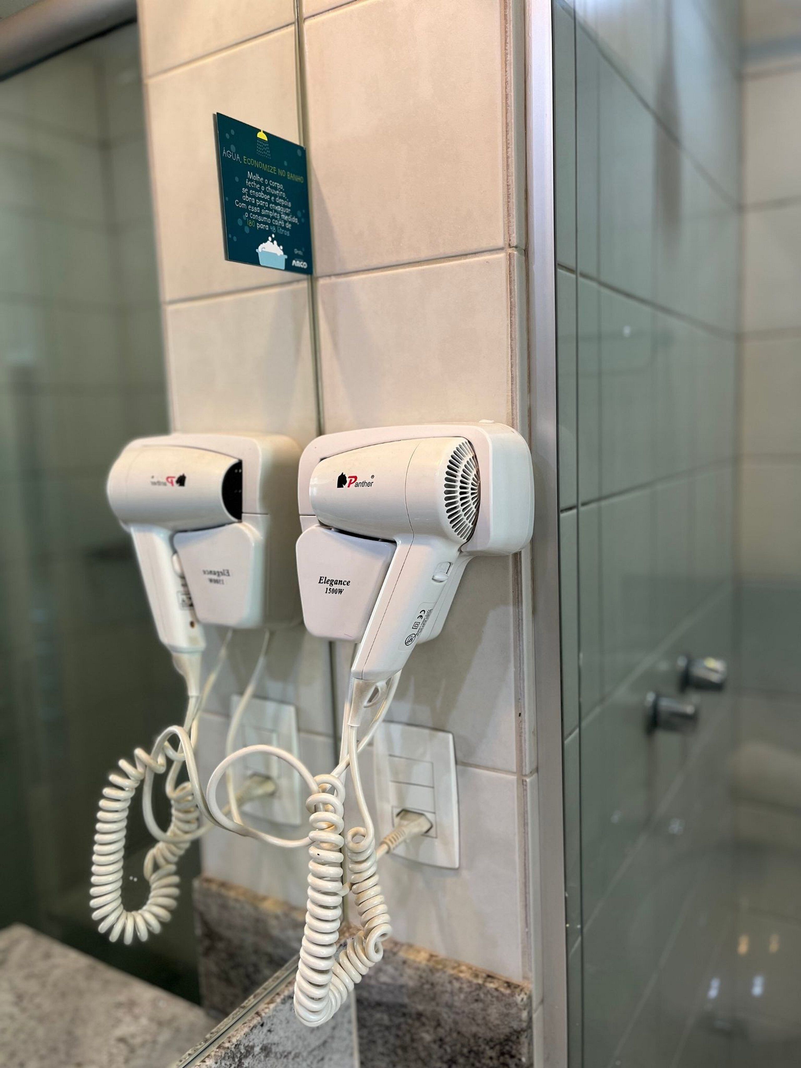 Uma casa de banho com dois secadores de cabelo brancos fixados na parede, um sinal com texto e um chão em azulejos.