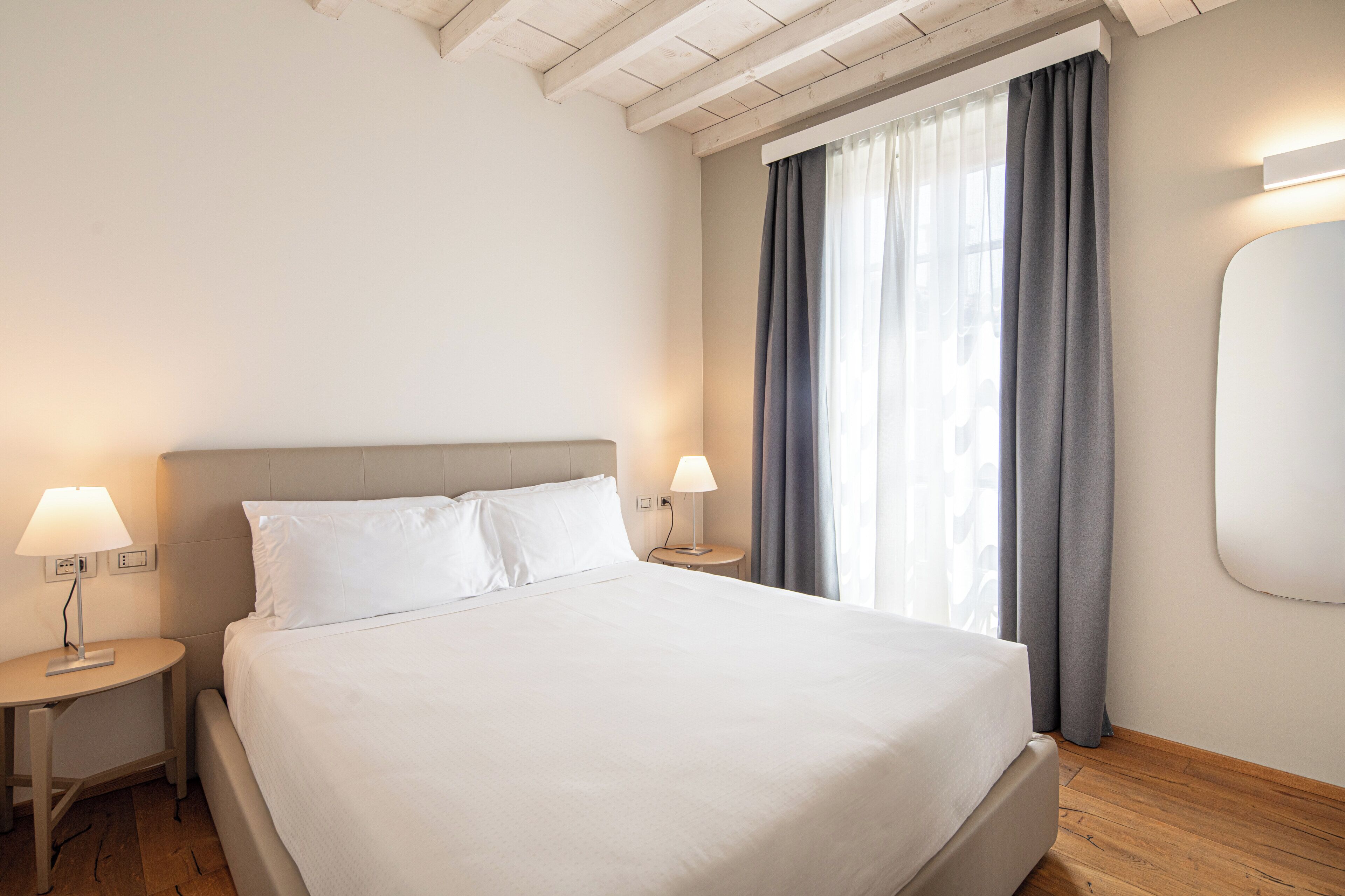 Cama bem arrumada com lençóis brancos, duas luminárias de cabeceira, uma janela com cortinas cinzas e piso de madeira.