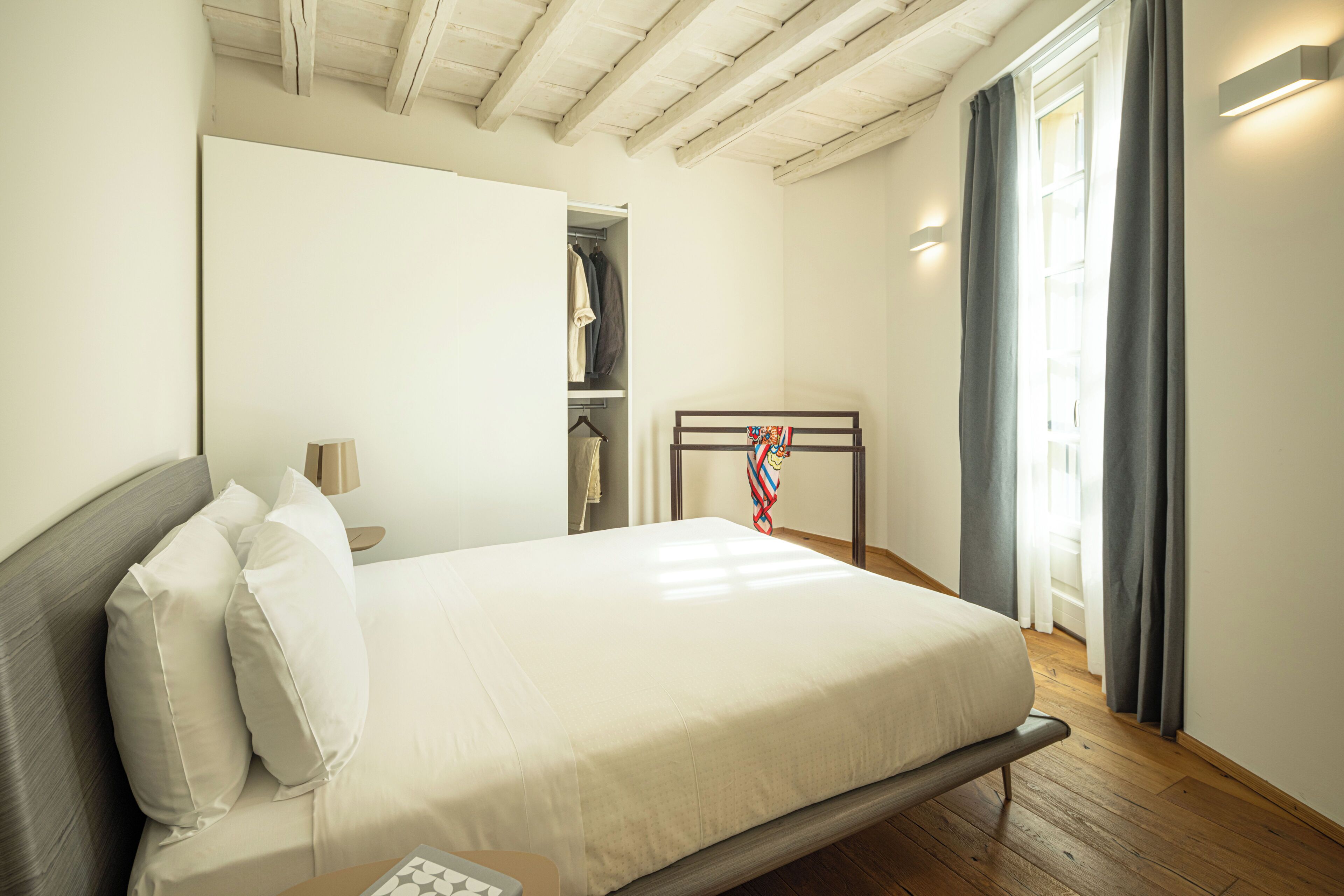 Quarto com cama, mesa de cabeceira, luminária, guarda-roupa e janela com cortinas.