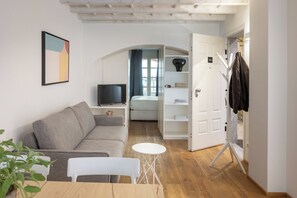 Uma sala de estar moderna com um sofá cinzento, uma televisão sobre um móvel, uma pequena mesa branca e uma porta que dá para outro cômodo.