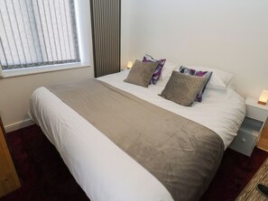 Uma cama bem arrumada com colcha e almofadas bege, uma janela com persianas e uma mesa de cabeceira com abajur.