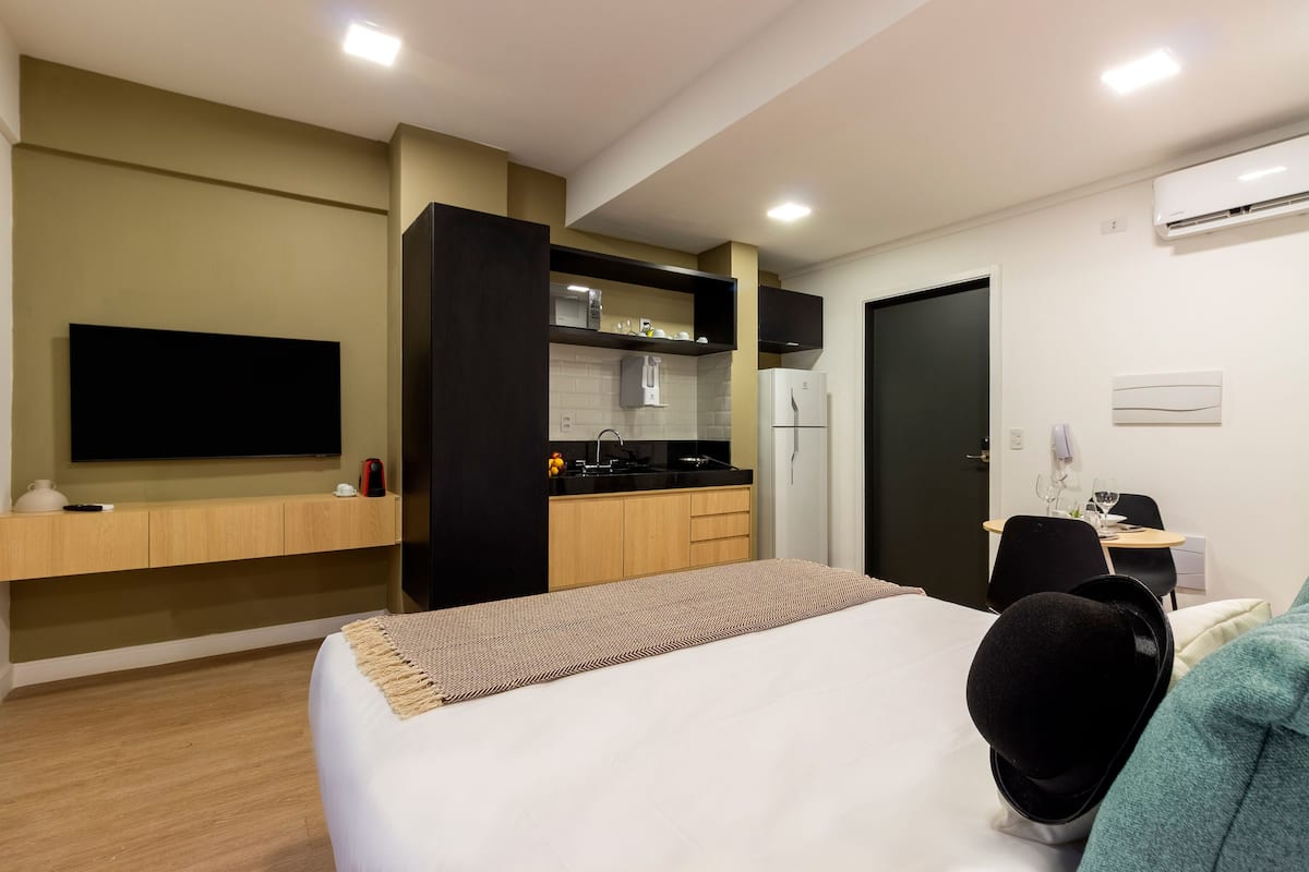 Quarto de hotel moderno com cama, televisão, cozinha compacta e área de refeições.
