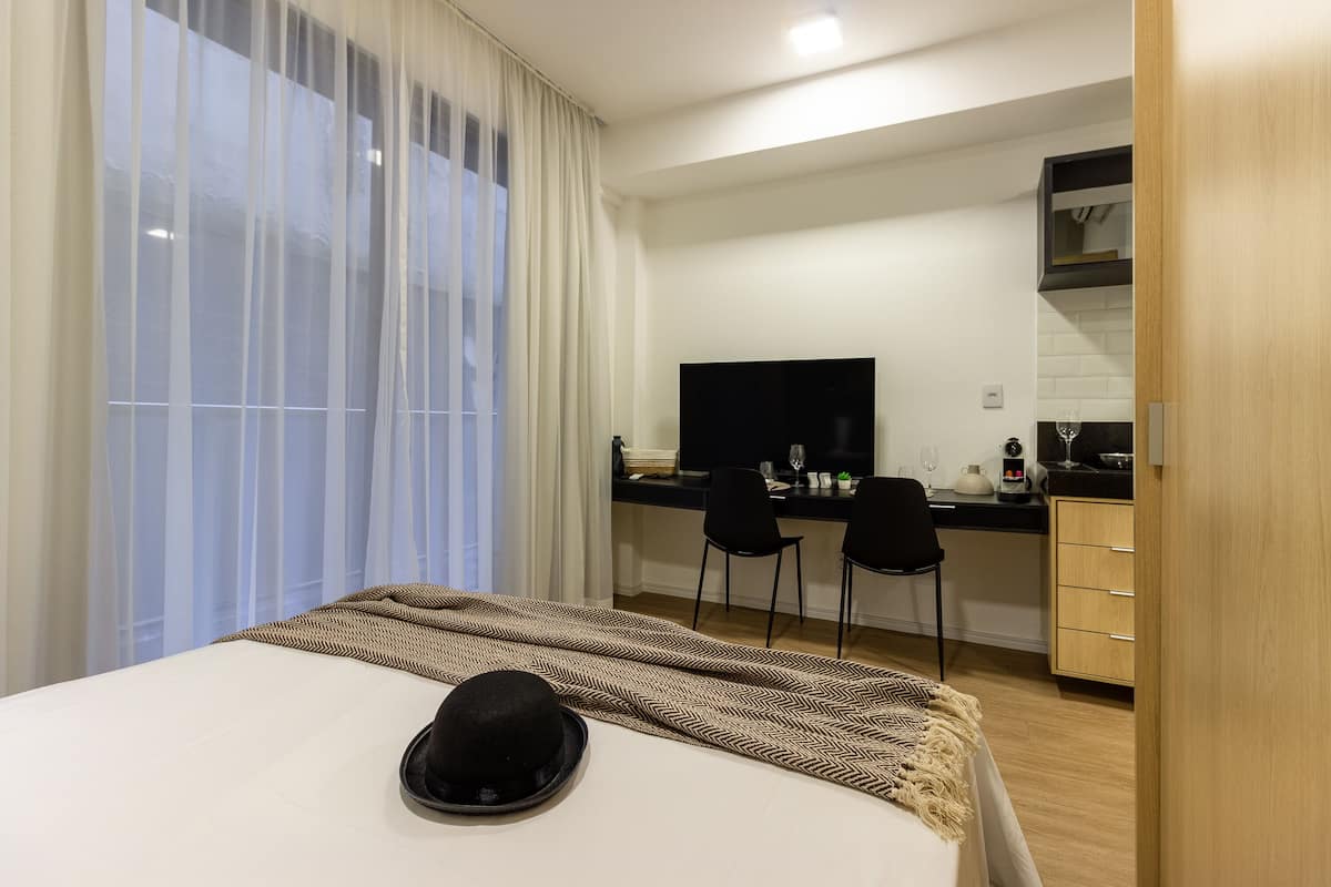 Quarto de hotel com cama, escrivaninha, duas cadeiras, televisão e cozinha compacta.