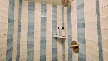 Área de duche com parede revestida a azulejos azuis e brancos, um chuveiro manual e duas garrafas de champô numa prateleira.