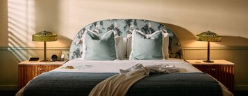 Cama bem arrumada com cabeceira floral, duas luminárias de cabeceira e mesa de cabeceira de madeira com relógio e telefone.