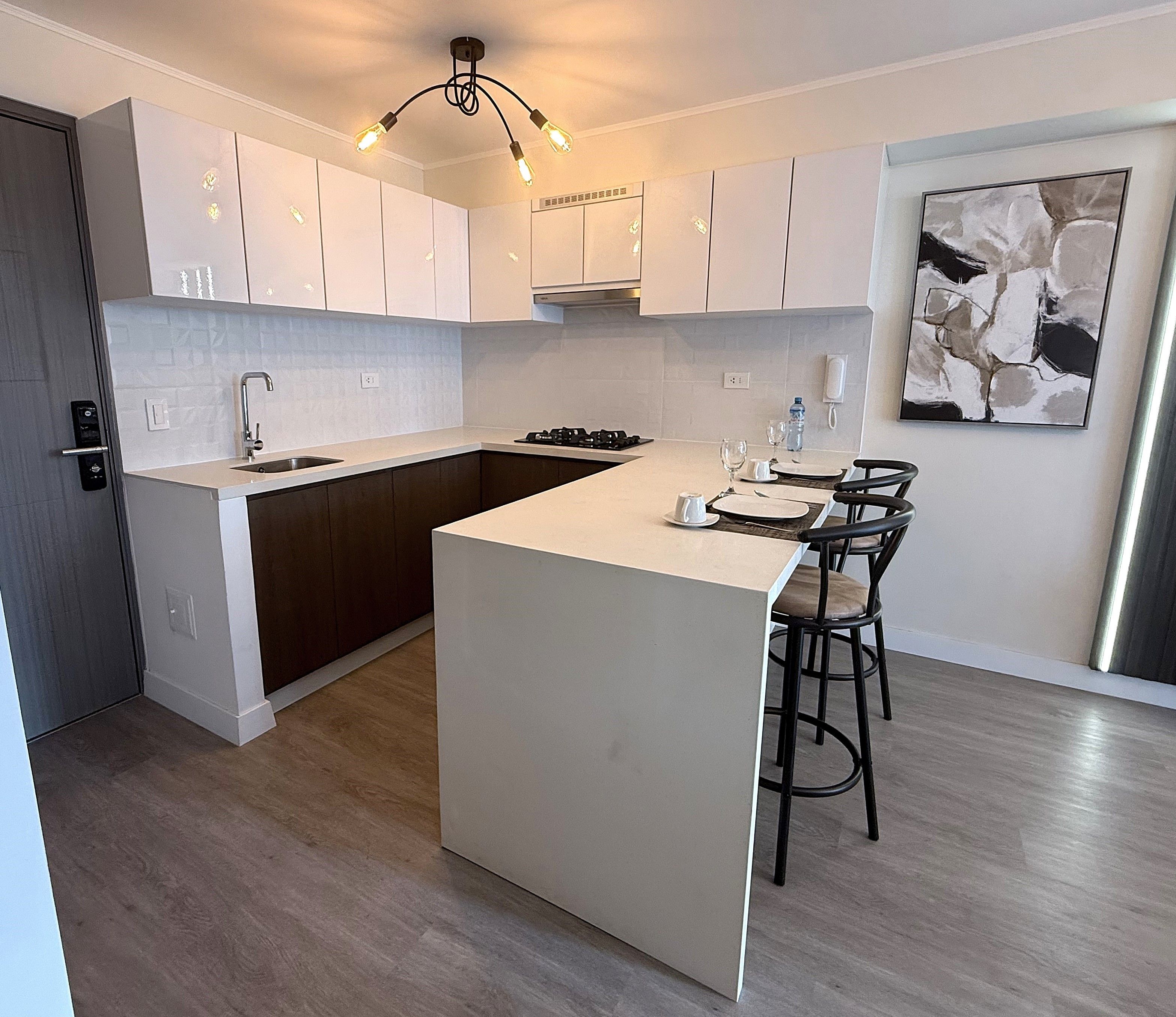 Cozinha moderna com armários brancos, uma ilha central, banquetas, fogão embutido e luminária pendente.