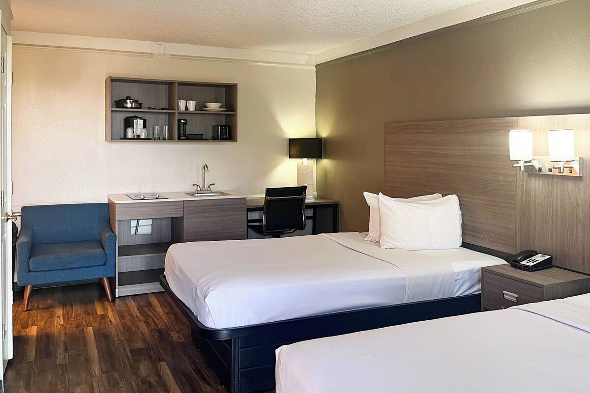 Quarto de hotel com duas camas, uma escrivaninha, uma cadeira, um pequeno kitchenette e telefone ao lado da cama.