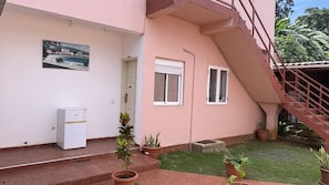 Um edifício rosa com escadas, porta e janela. Há plantas em vasos e uma pequena geladeira no chão.