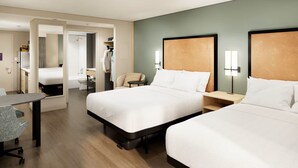 Quarto de hotel com duas camas, uma escrivaninha, uma cadeira, uma mesinha e banheiro visível por uma porta aberta.