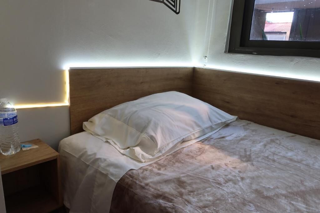 Quarto de hotel pequeno com uma cama de solteiro, mesa de cabeceira, uma garrafa de água e janela com vista para prédios.