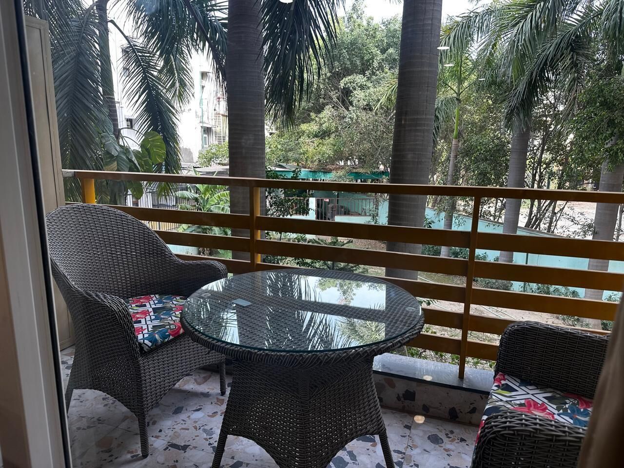 Uma varanda com móveis de vime, uma mesa de vidro e vista para palmeiras e edifícios.