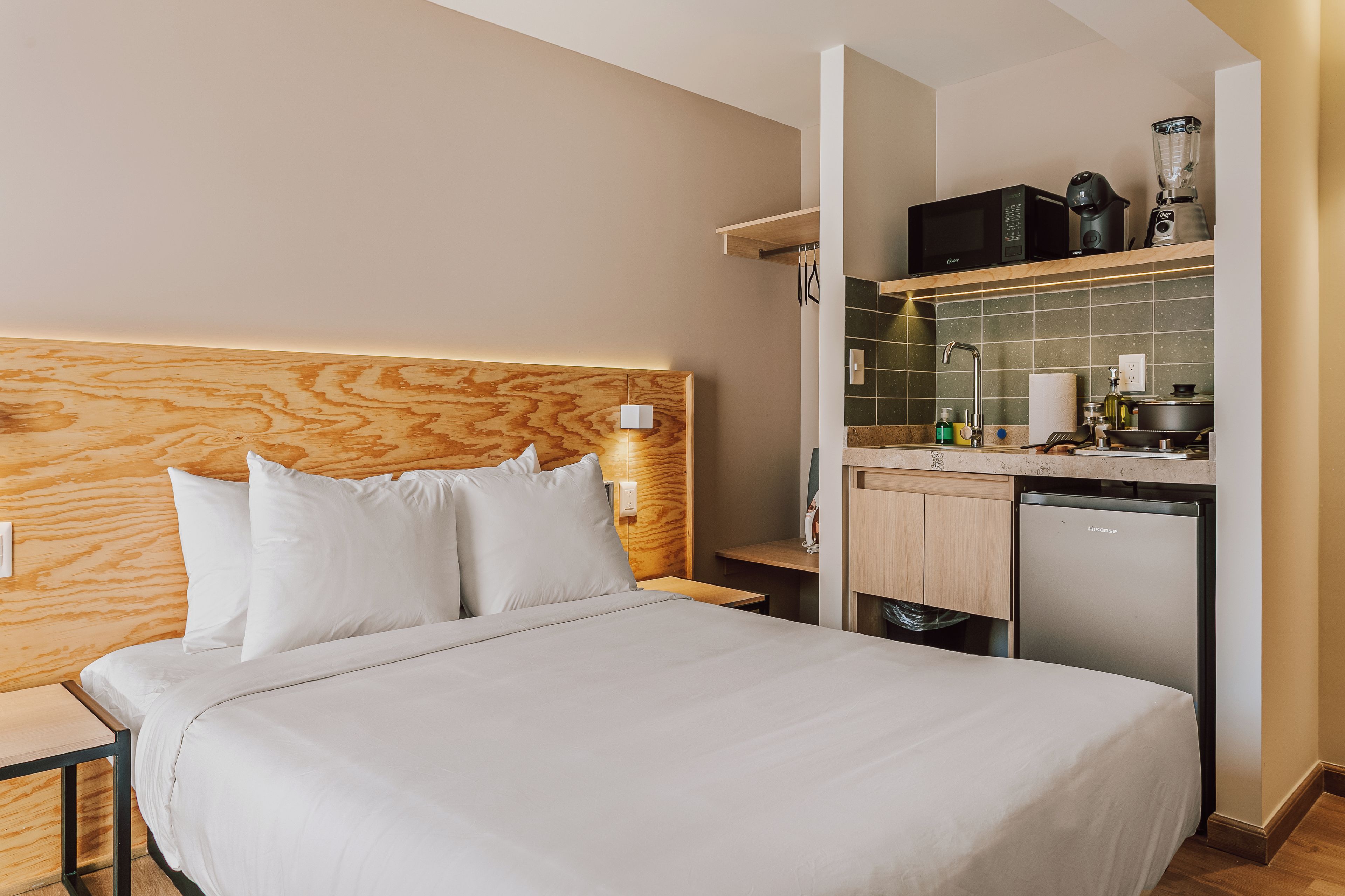 Quarto de hotel com cabeceira de madeira, cama com roupa de cama branca, e uma pequena cozinha equipada com micro-ondas, liquidificador e geladeira.