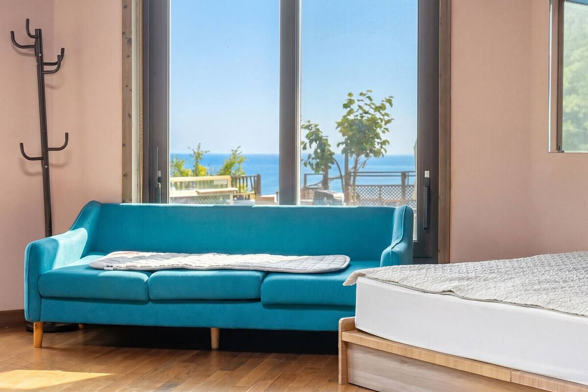 Quarto com sofá azul, cama, cabideiro e vista para o mar.