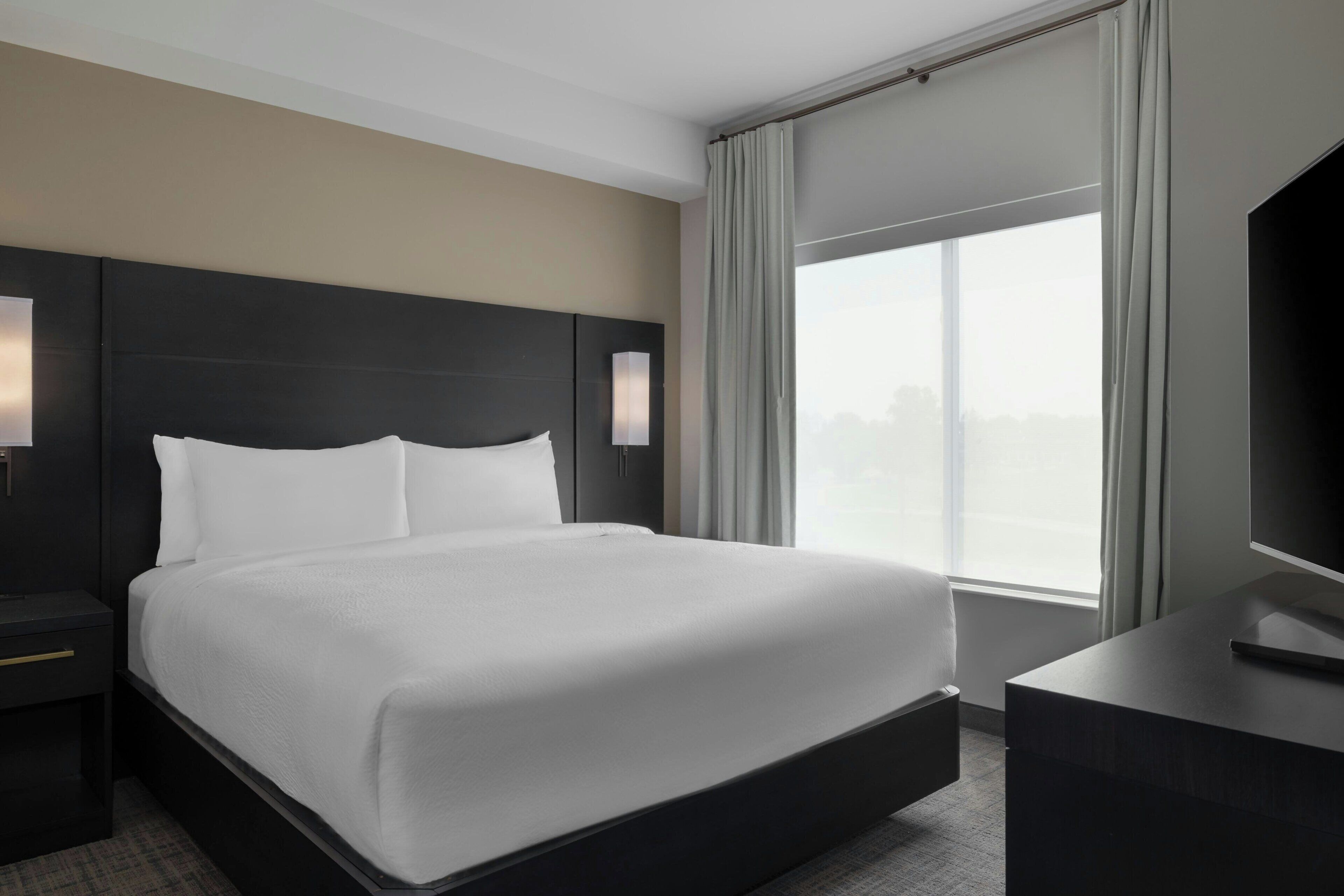 Quarto de hotel com uma cama grande, televisão de tela plana, mesa de cabeceira e janela com cortinas.