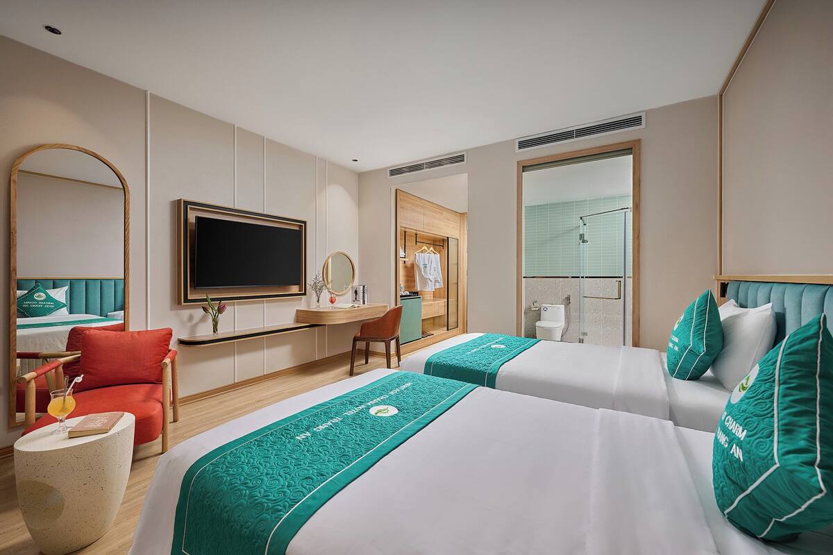Quarto de hotel com duas camas, televisão, escrivaninha, cadeira e banheiro visível por uma porta aberta.