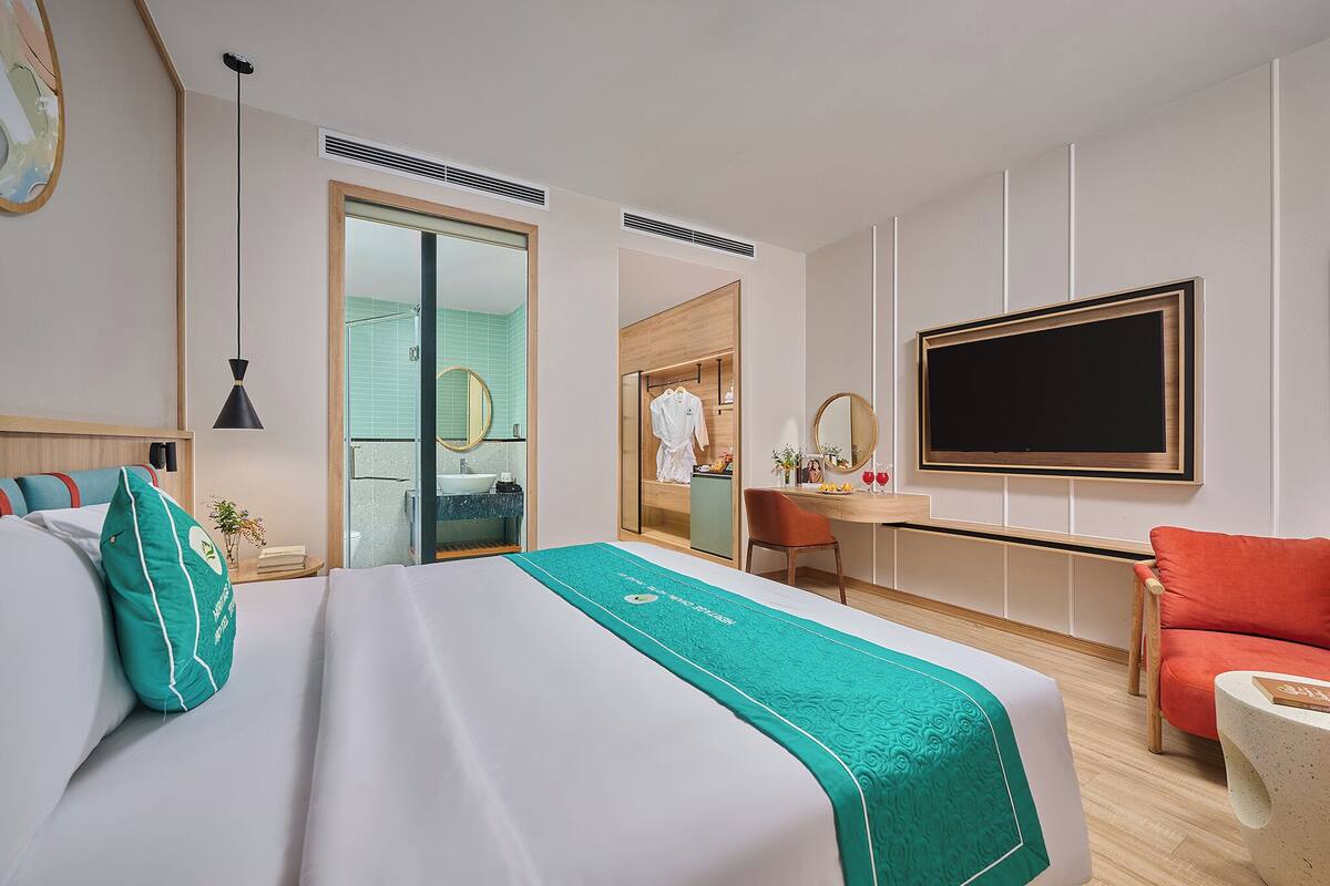 Quarto de hotel com cama, escrivaninha, cadeira, televisão e banheiro visível através de uma porta aberta.