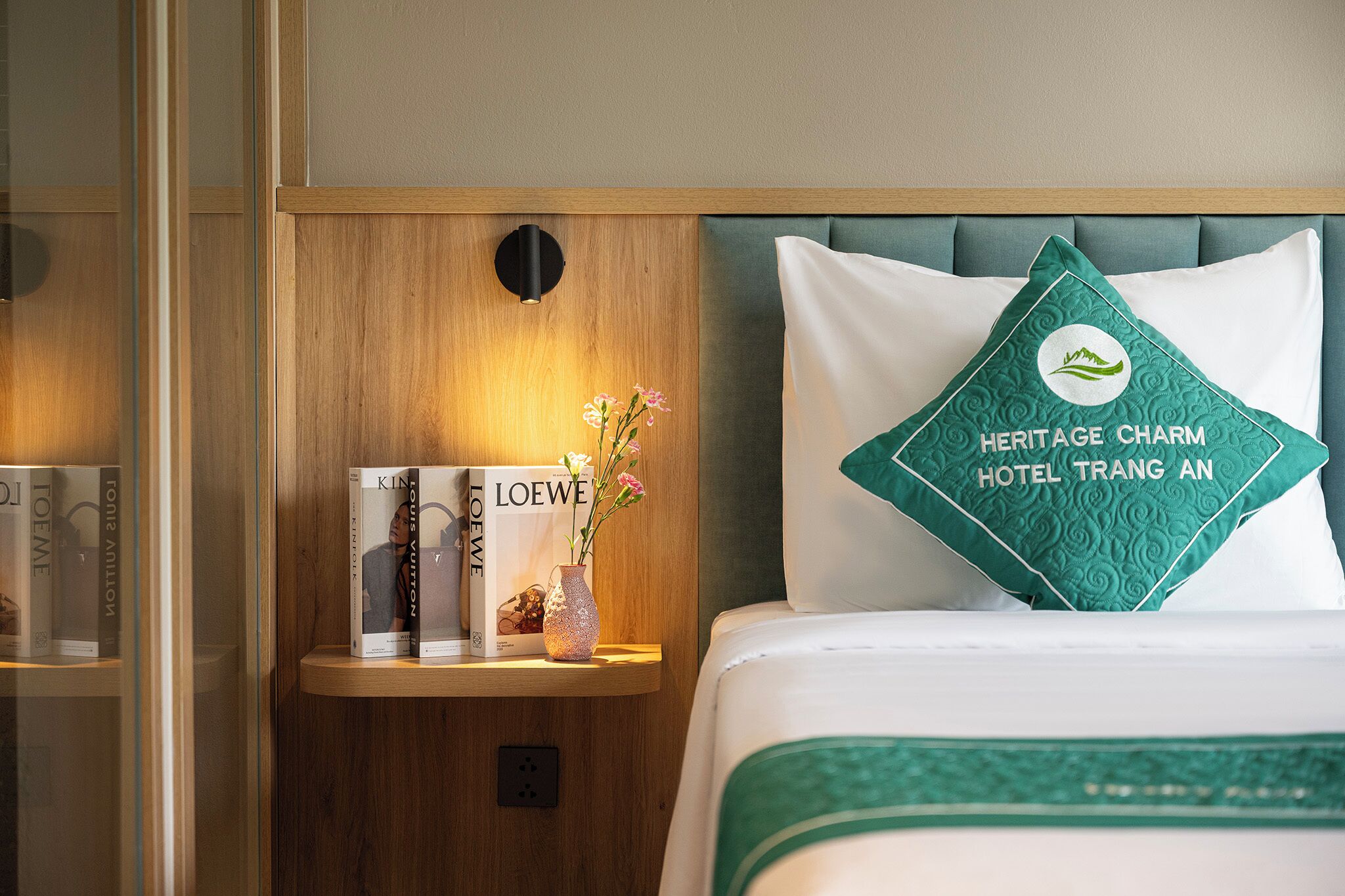 Quarto de hotel com cama, mesa de cabeceira com livros e um vaso, e travesseiro com o nome do hotel.