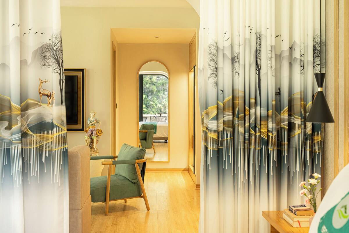 Quarto com uma cadeira verde, uma mesinha com livros e um vaso, e uma janela com vista para vegetação.