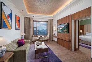 Quarto de hotel moderno com sofá, mesa de centro, televisão e uma grande janela com vista para as montanhas.
