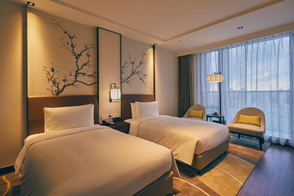 Quarto de hotel com duas camas, uma mesa, uma cadeira e janelas amplas com cortinas.