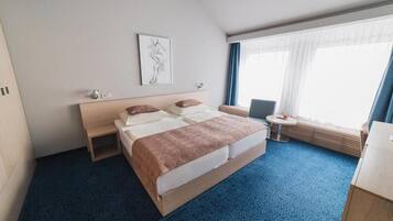 Quarto de hotel com uma cama, mesas de cabeceira, uma pequena mesa redonda, uma cadeira, uma janela com cortinas e uma obra de arte na parede.