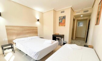 Quarto de hotel com duas camas, uma escrivaninha, uma cadeira, um quadro na parede e uma porta que dá para outro cômodo.