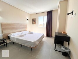 Quarto de hotel com cama, escrivaninha, cadeira, lixeira e janela com cortinas.