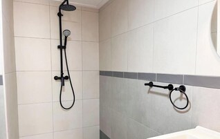 Casa de banho com azulejos brancos, chuveiro com mangueira preta e um suporte para toalhas.