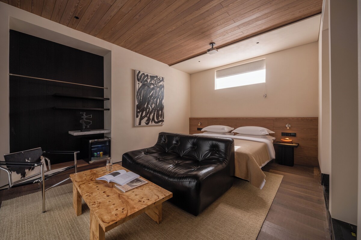 Quarto moderno com sofá de couro preto, mesa de centro de madeira, cadeira de metal preto, cama com roupa de cama branca, mesa de cabeceira com abajur e uma grande pintura abstrata na parede.