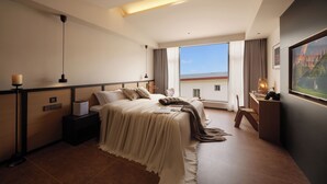 Quarto de hotel moderno com uma cama grande, uma escrivaninha, uma cadeira, uma mesa de cabeceira, um abajur e um quadro na parede.