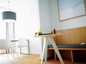 Quarto moderno com um banco de madeira, uma mesa redonda com uma garrafa e frutas, e uma luminária pendente.