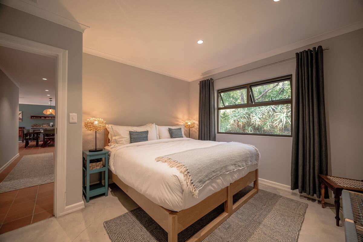 Quarto com cama, mesas de cabeceira, janela com cortinas e vista para o verde lá fora.