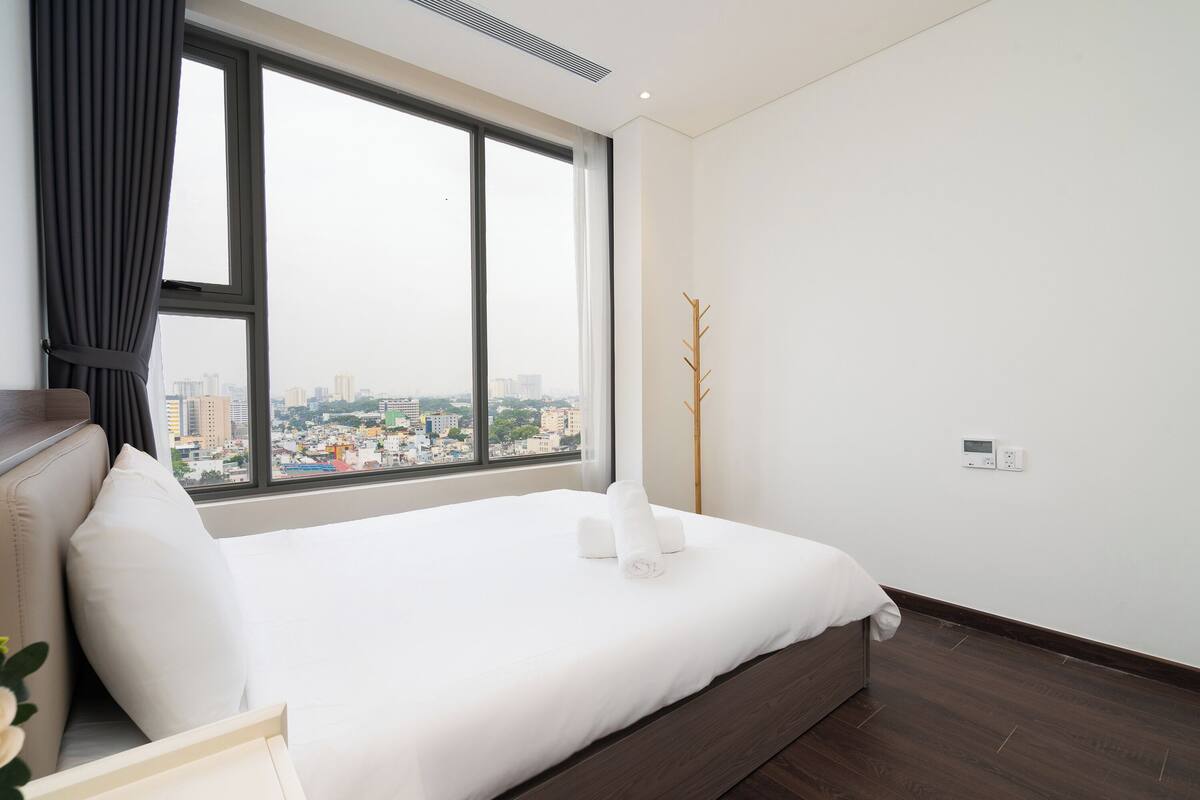 Quarto de hotel com uma janela grande, uma cama, uma mesinha de cabeceira e um termostato fixado na parede.