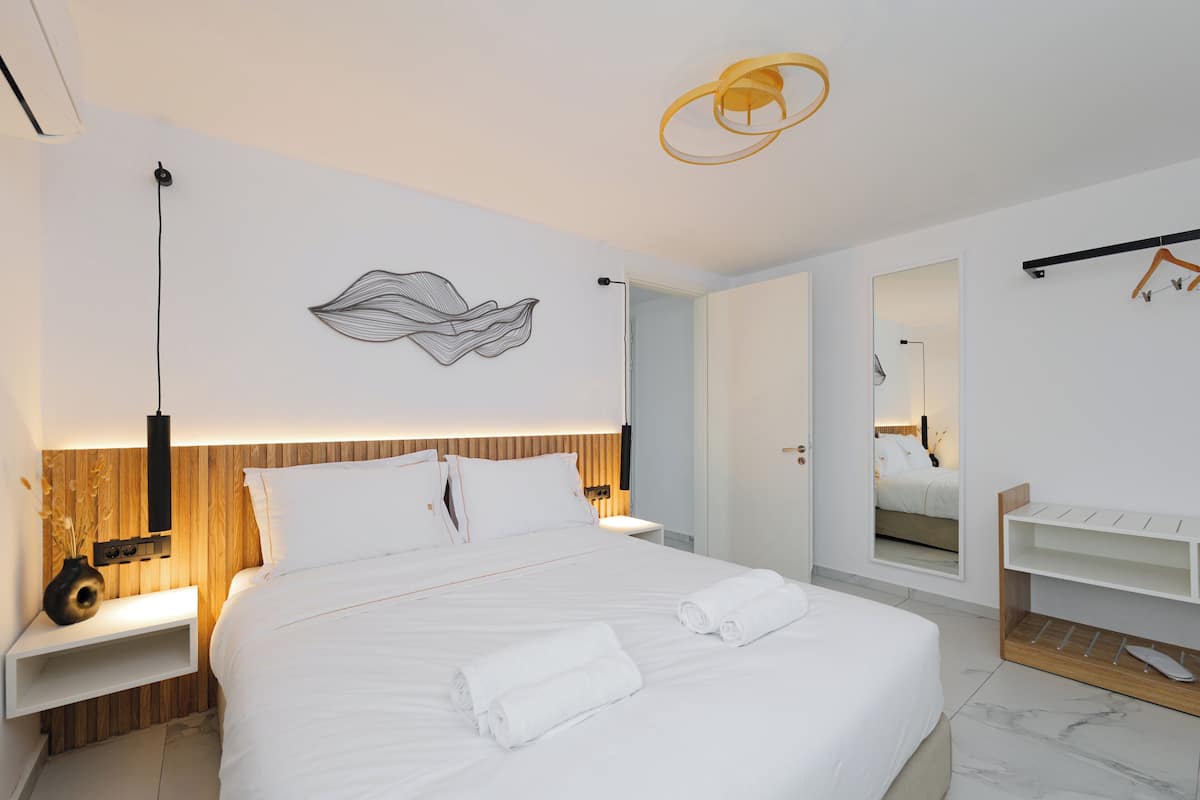 Quarto de hotel moderno com uma cama grande, uma mesa de cabeceira, uma obra de arte na parede, um espelho e uma estante de madeira.