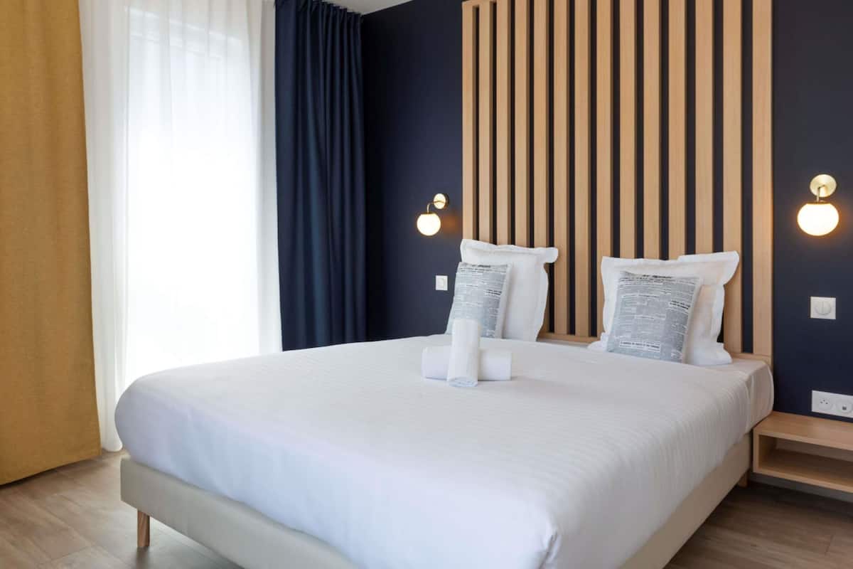 Quarto de hotel moderno com uma cama grande, cabeceira de madeira e duas luminárias fixadas na parede.