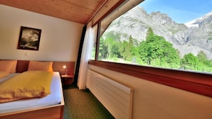 Quarto com cama, mesa de cabeceira, janela com vista para as montanhas e quadro na parede.