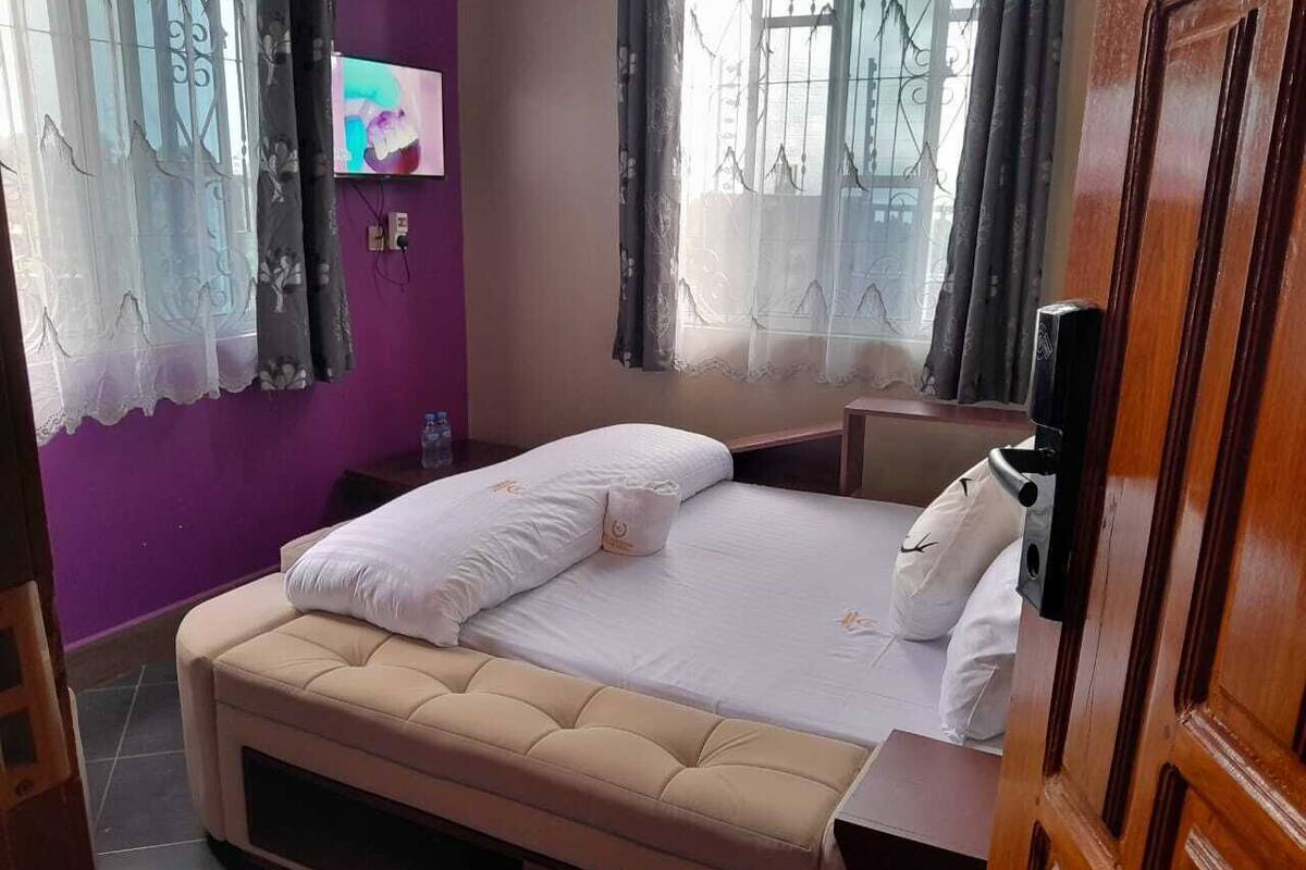 Quarto com cama, mesa de cabeceira, televisão e janela com cortinas.