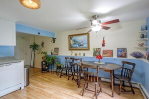 Uma sala de jantar com mesa redonda, cadeiras de madeira, ventilador de teto e diversos objetos decorativos nas paredes, incluindo um quadro emoldurado e um leque.