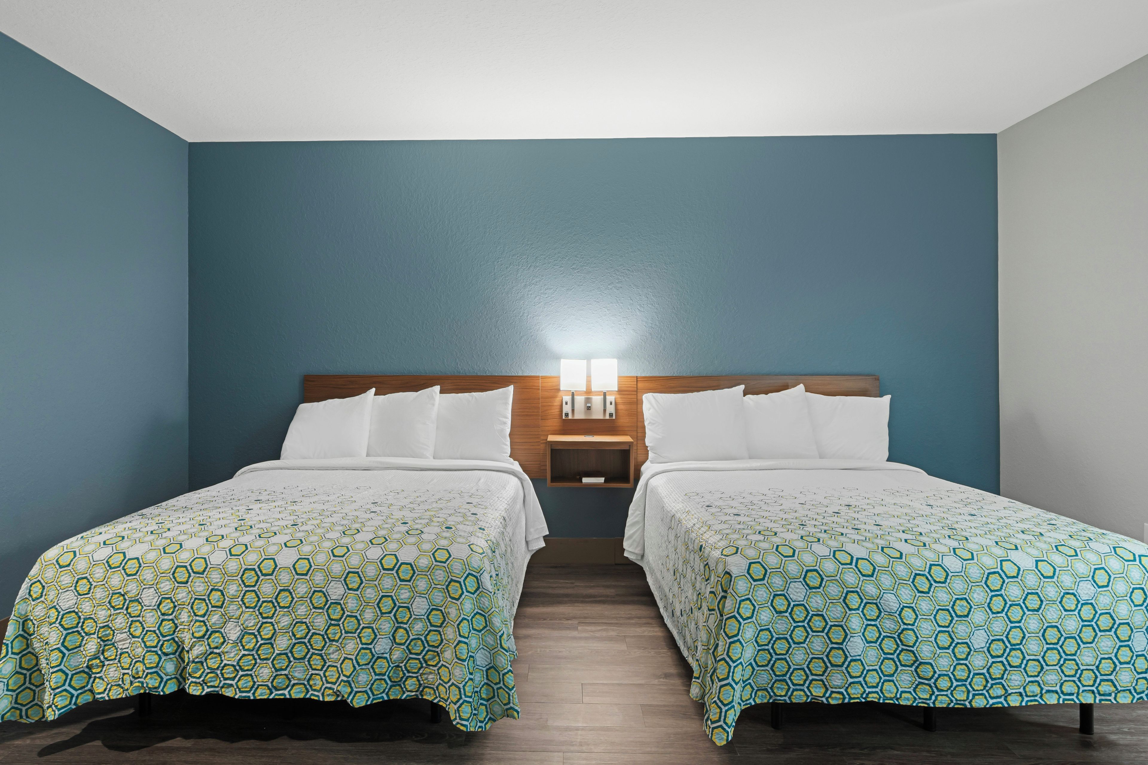 Quarto de hotel com duas camas, cabeceira de madeira, mesa de cabeceira com abajur e parede azul.