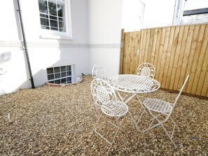 Um pátio com uma mesa e cadeiras de metal branco, cerca de madeira e um edifício branco com uma janela.