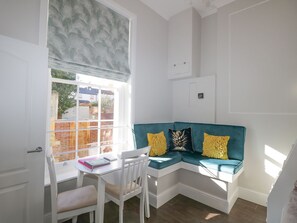 Quarto nos fundos com janela, mesa branca com cadeira e sofá azul-turquesa com almofadas amarelas.