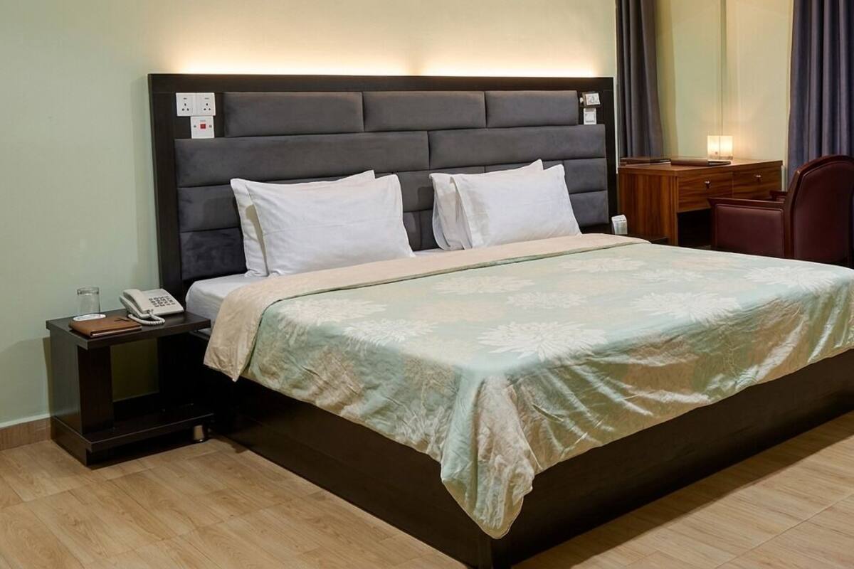 Quarto de hotel com cama, mesa de cabeceira, luminária e escrivaninha com cadeira.