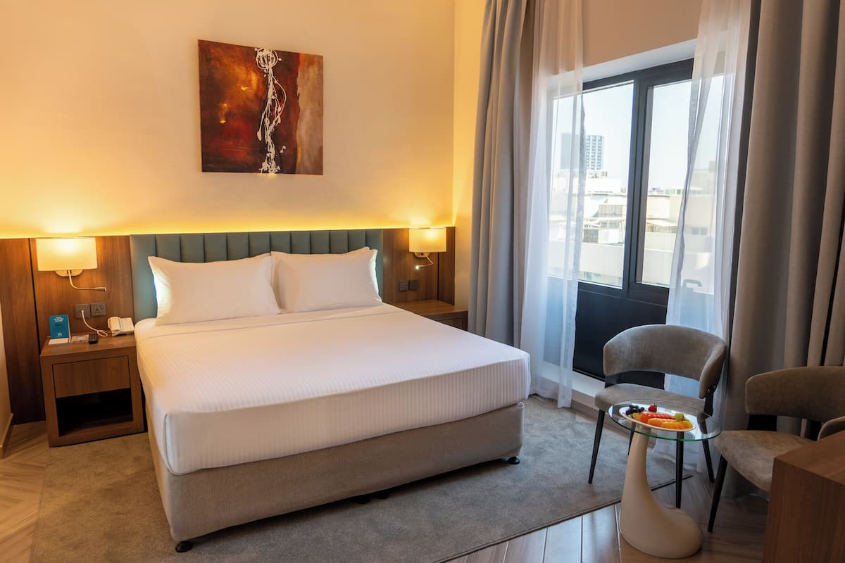 Quarto de hotel com uma cama grande, luminárias laterais, uma mesinha com frutas, uma cadeira e vista para a cidade pela janela.