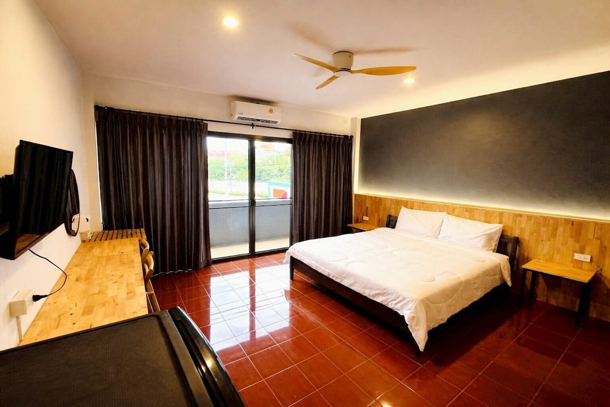 Quarto de hotel com cama, escrivaninha, televisão, ventoinha no teto e vista através de uma janela grande.