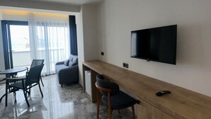 Uma sala de estar moderna com televisão de tela plana na parede, uma escrivaninha de madeira, uma mesa de jantar com cadeiras e um sofá.