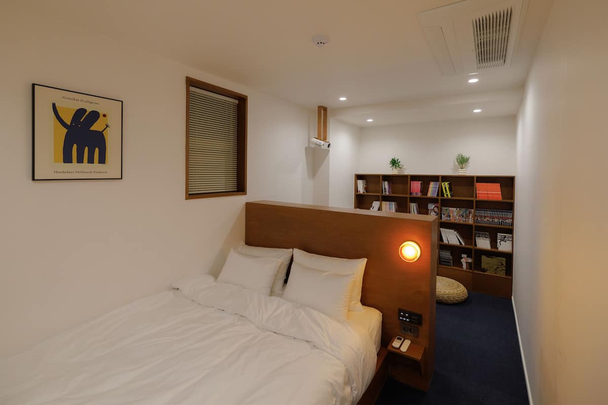 Uma cama bem arrumada com lençóis brancos, uma mesa de cabeceira com abajur, uma estante com livros e objetos decorativos, e um quadro na parede.