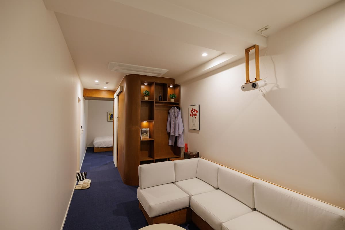 Quarto de hotel com sofá branco, armário de madeira, cama pequena e corredor que dá para outro quarto.