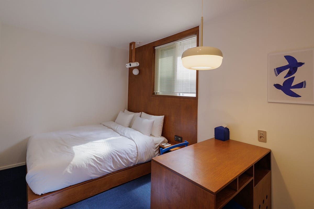 Quarto com cama de madeira, escrivaninha, janela com persianas, luminária na parede e uma obra de arte emoldurada.