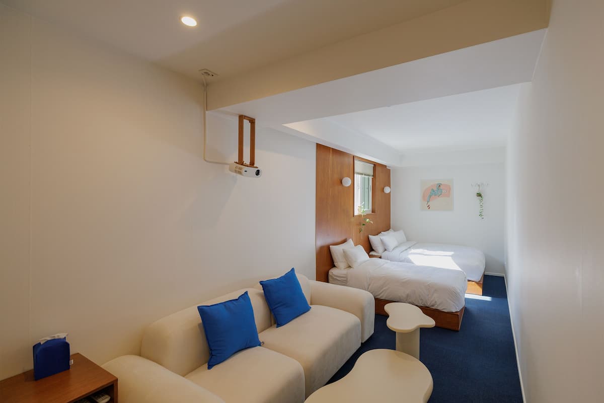 Quarto de hotel com sofá, mesa pequena, cama e projetor instalado no teto.